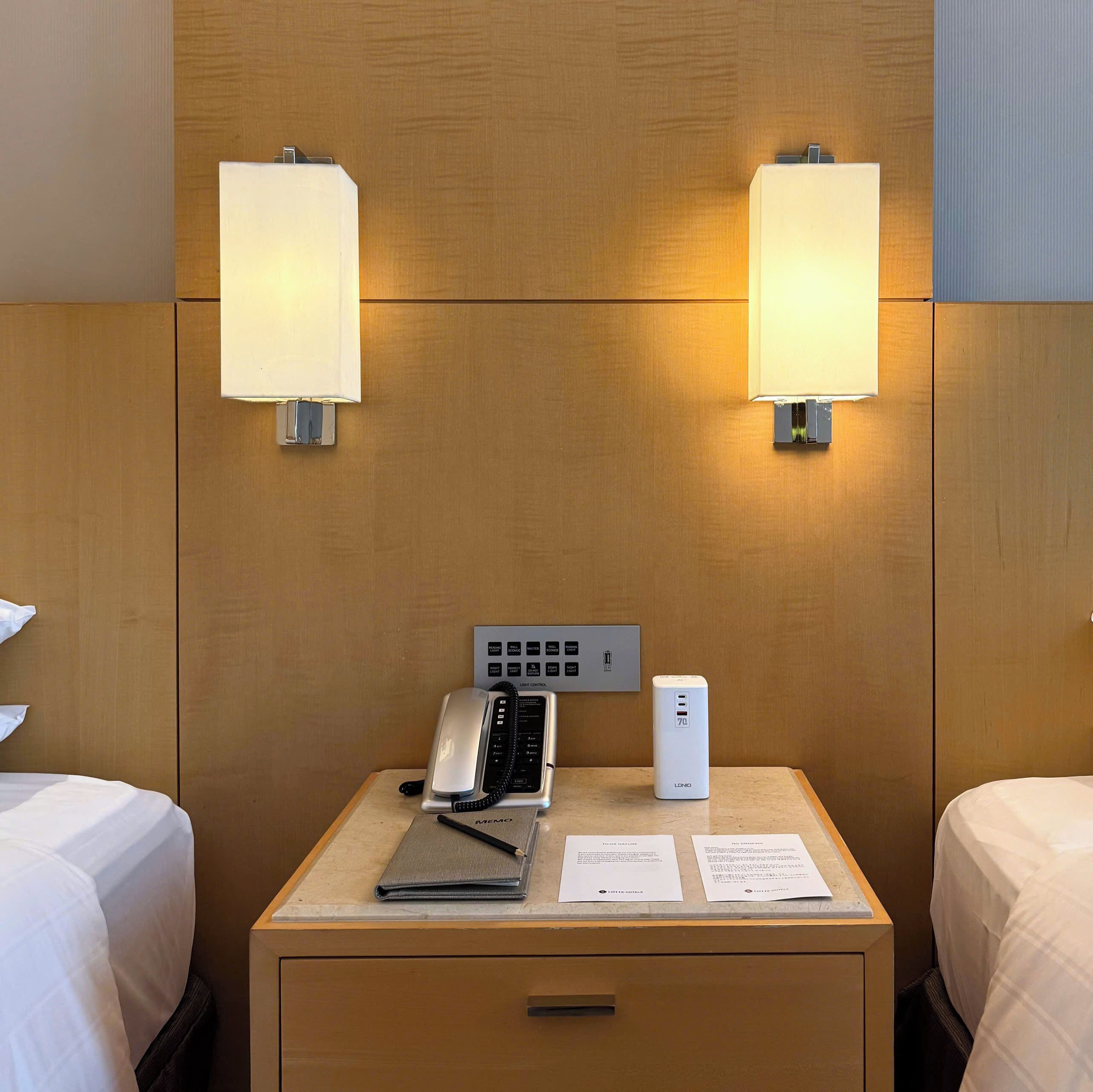 Quarto de hotel com cabeceira de madeira, mesa de cabeceira com telefone, régua de tomada e duas luminárias.