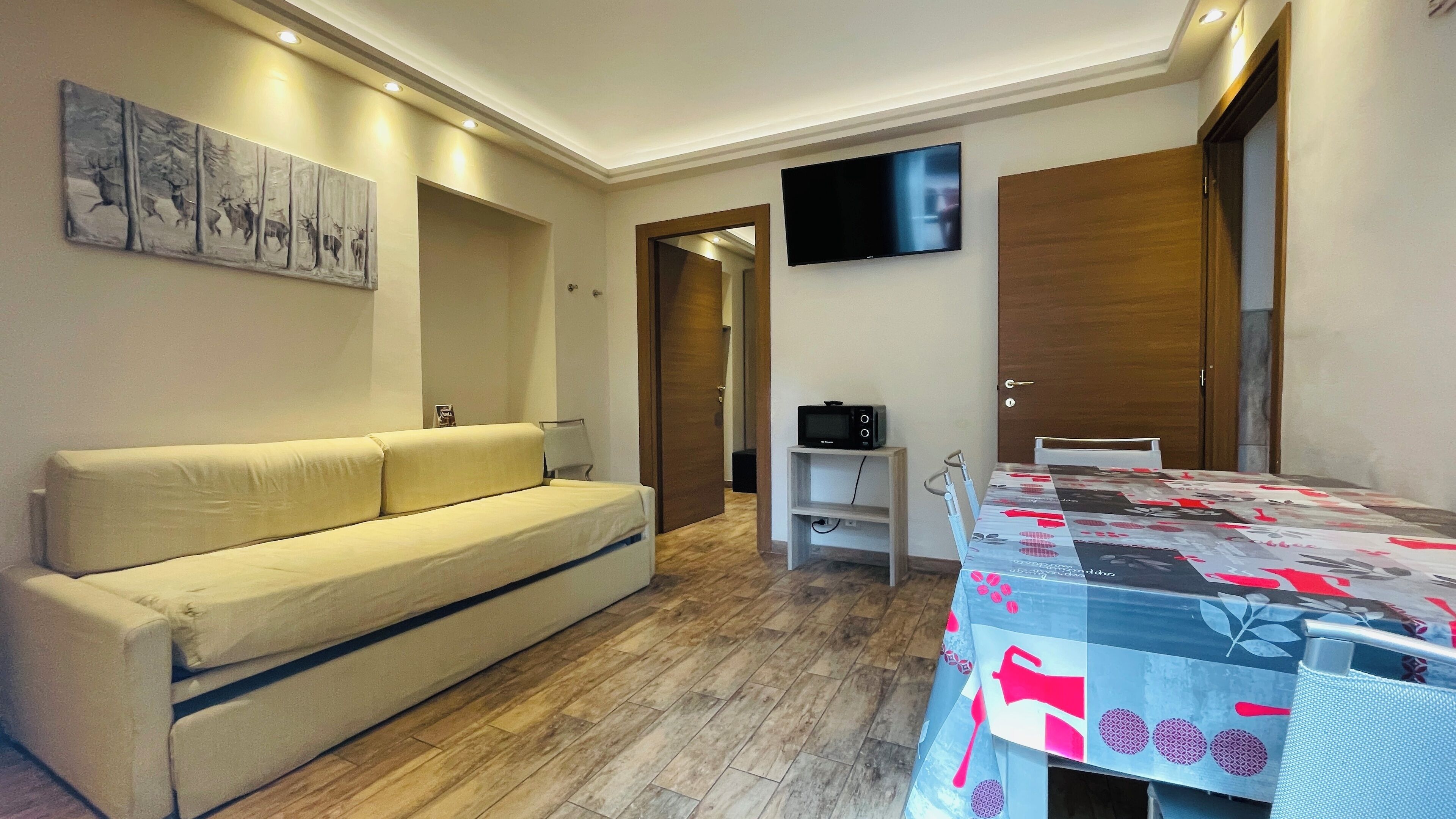 Uma sala de estar com um sofá, uma TV fixada na parede e uma mesa com toalha.