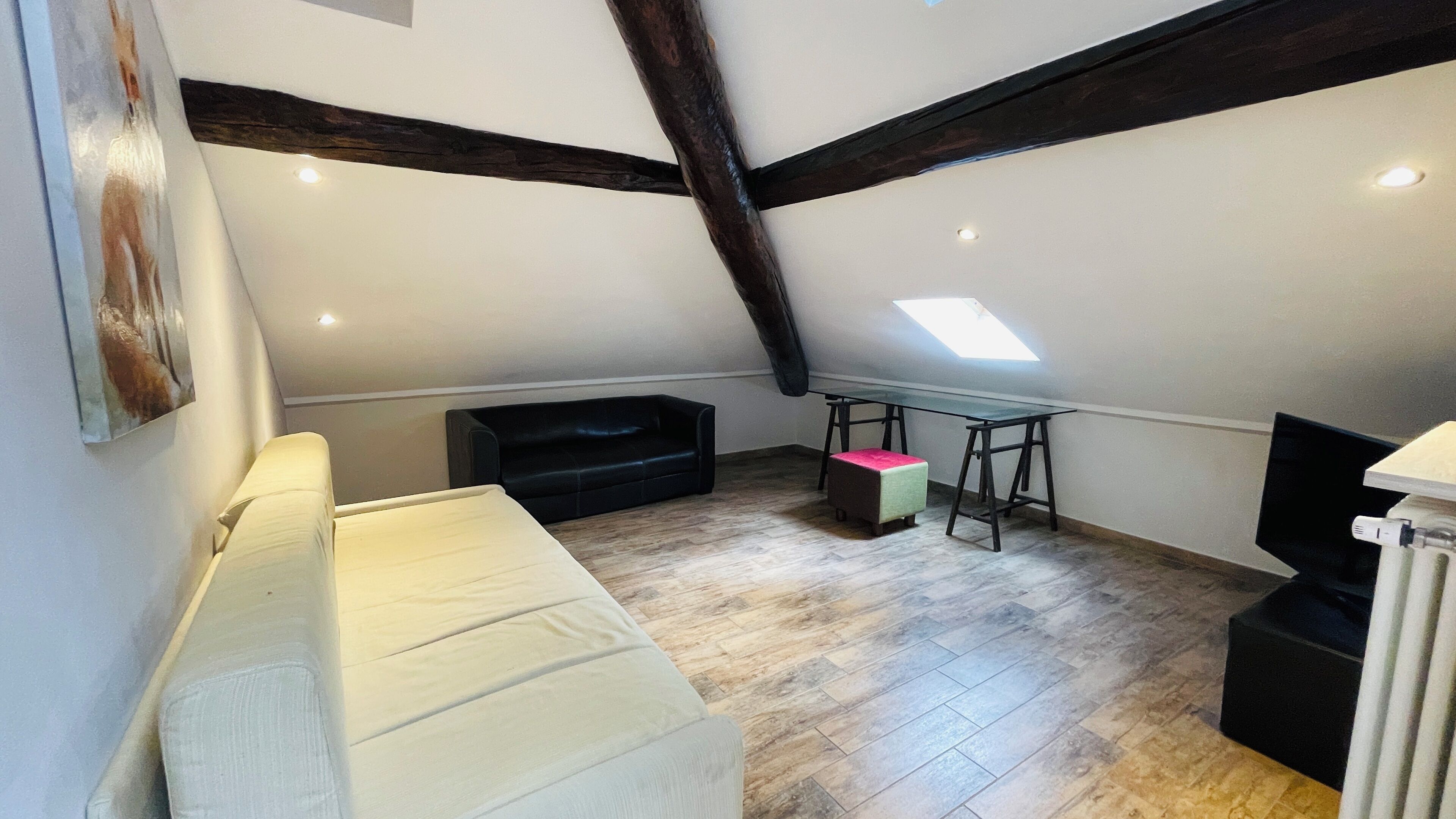 Um quarto no estilo loft com teto inclinado, vigas de madeira aparentes, um sofá branco, um sofá de couro preto, uma mesinha e um radiador.