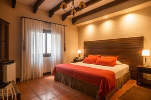 Quarto com cama grande, cabeceira de madeira, duas mesas de cabeceira com luminárias, um criado-mudo e um banco, além de uma janela com cortinas.