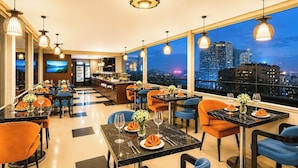 Um restaurante moderno com vista para a cidade, com mesas de mármore preto, cadeiras laranja e azuis e iluminação pendente.
