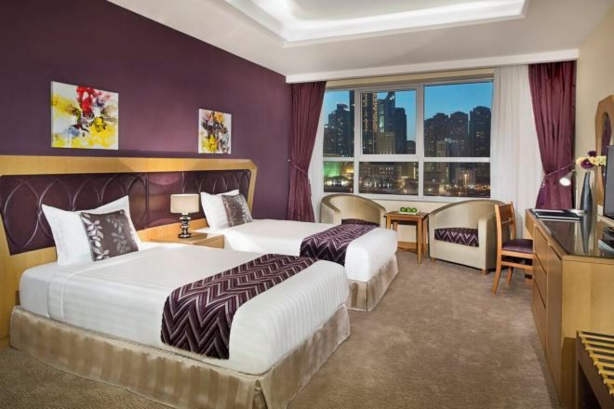 Quarto de hotel com duas camas, uma escrivaninha, uma cadeira e vista para a paisagem urbana.