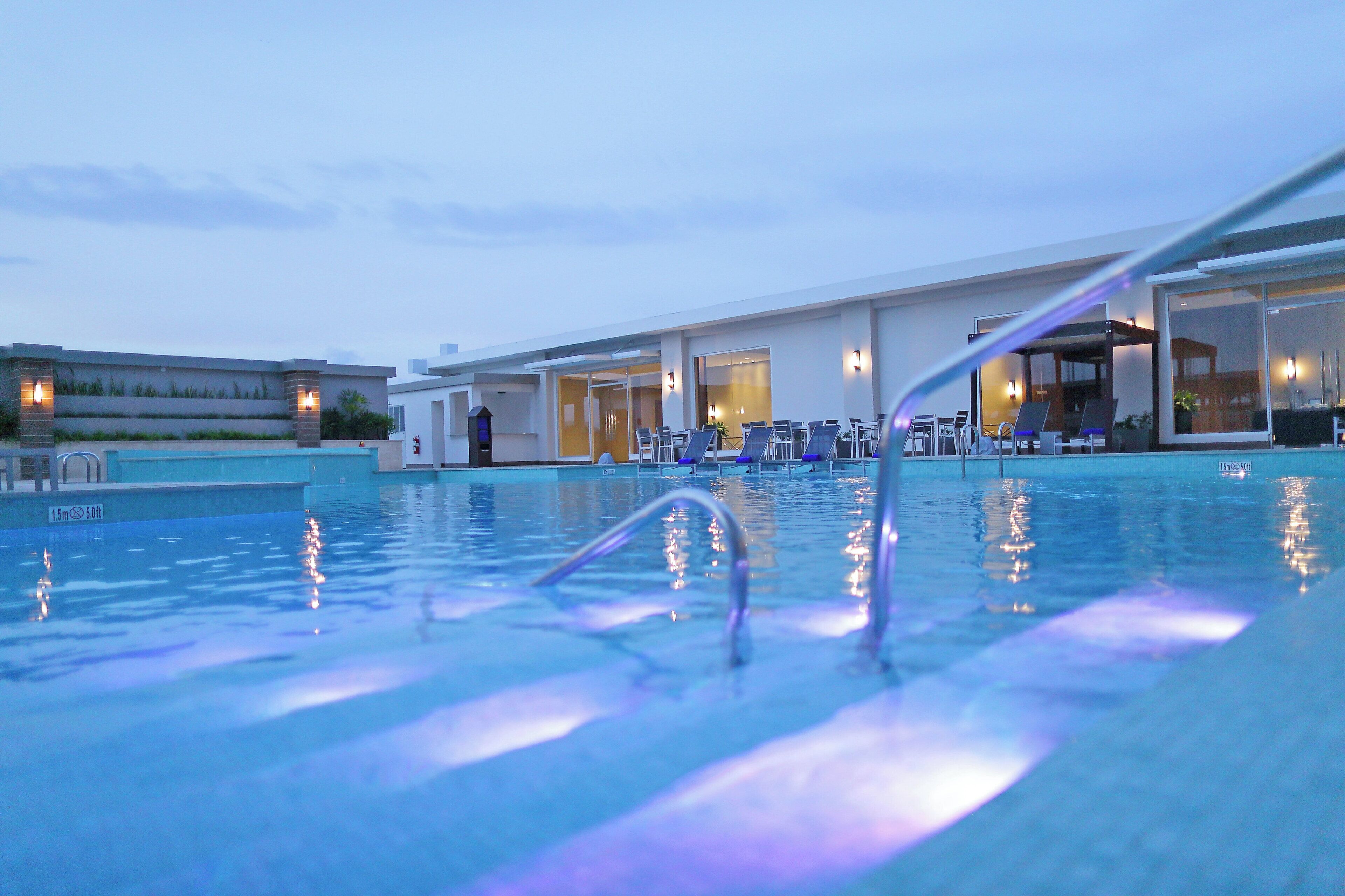 Piscina com superfície de água límpida, área ao redor da piscina com espreguiçadeiras e um edifício com amplas janelas e portas.