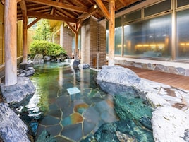 Área de banho onsen tradicional japonesa, com vigas de madeira, paredes de pedra e vista para o exterior.