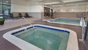 Piscina interior com borda azulejada, área de estar com cadeiras e vista clara pelas janelas.