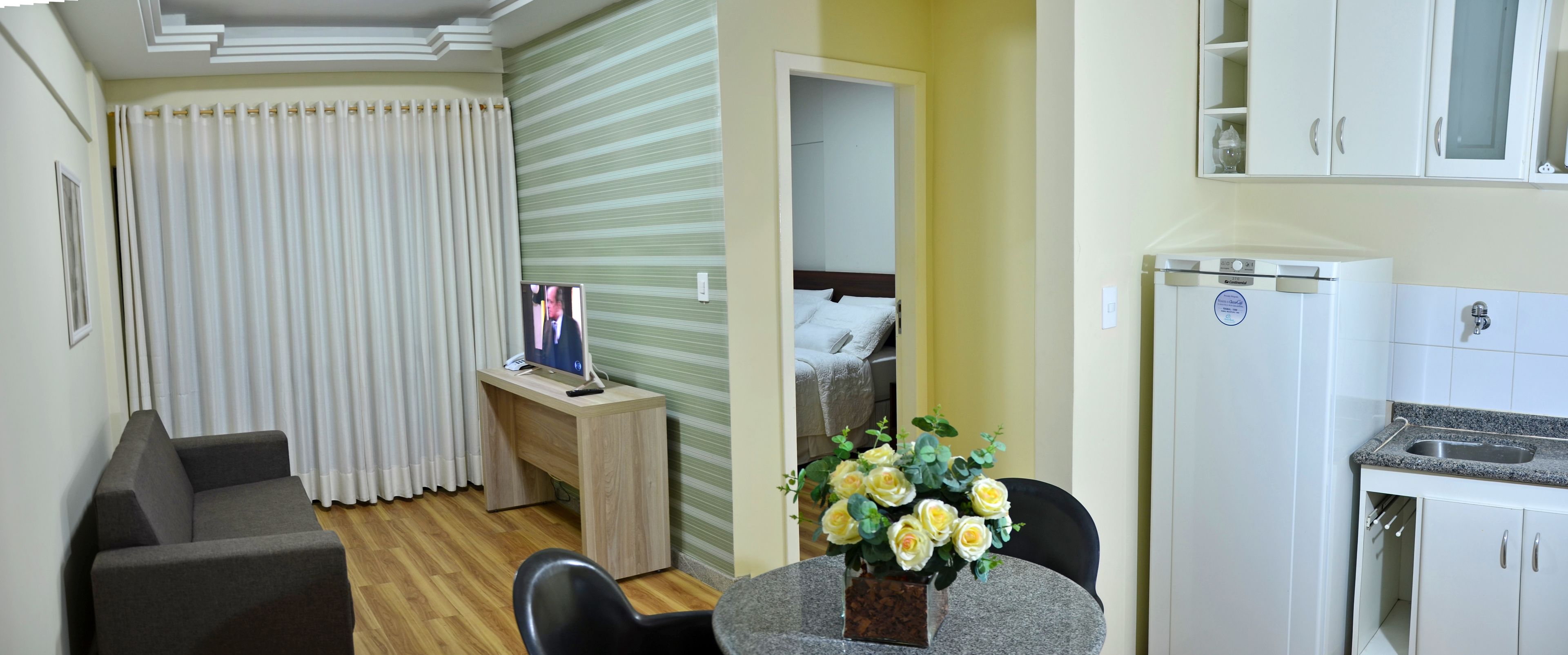 Quarto de hotel pequeno e funcional, com sofá, mesa de jantar com um vaso de flores, cozinha compacta e banheiro visível por uma porta aberta.