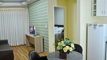 Quarto de hotel pequeno e funcional, com sofá, mesa de jantar com um vaso de flores, cozinha compacta e banheiro visível por uma porta aberta.