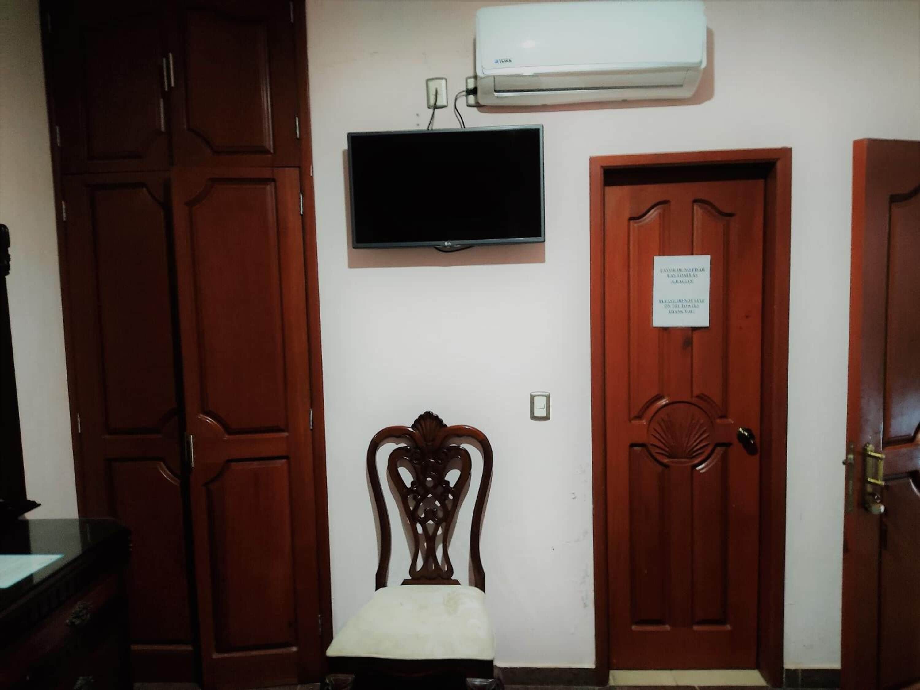Quarto com guarda-roupa de madeira, televisão na parede, uma cadeira de madeira e porta com aviso.
