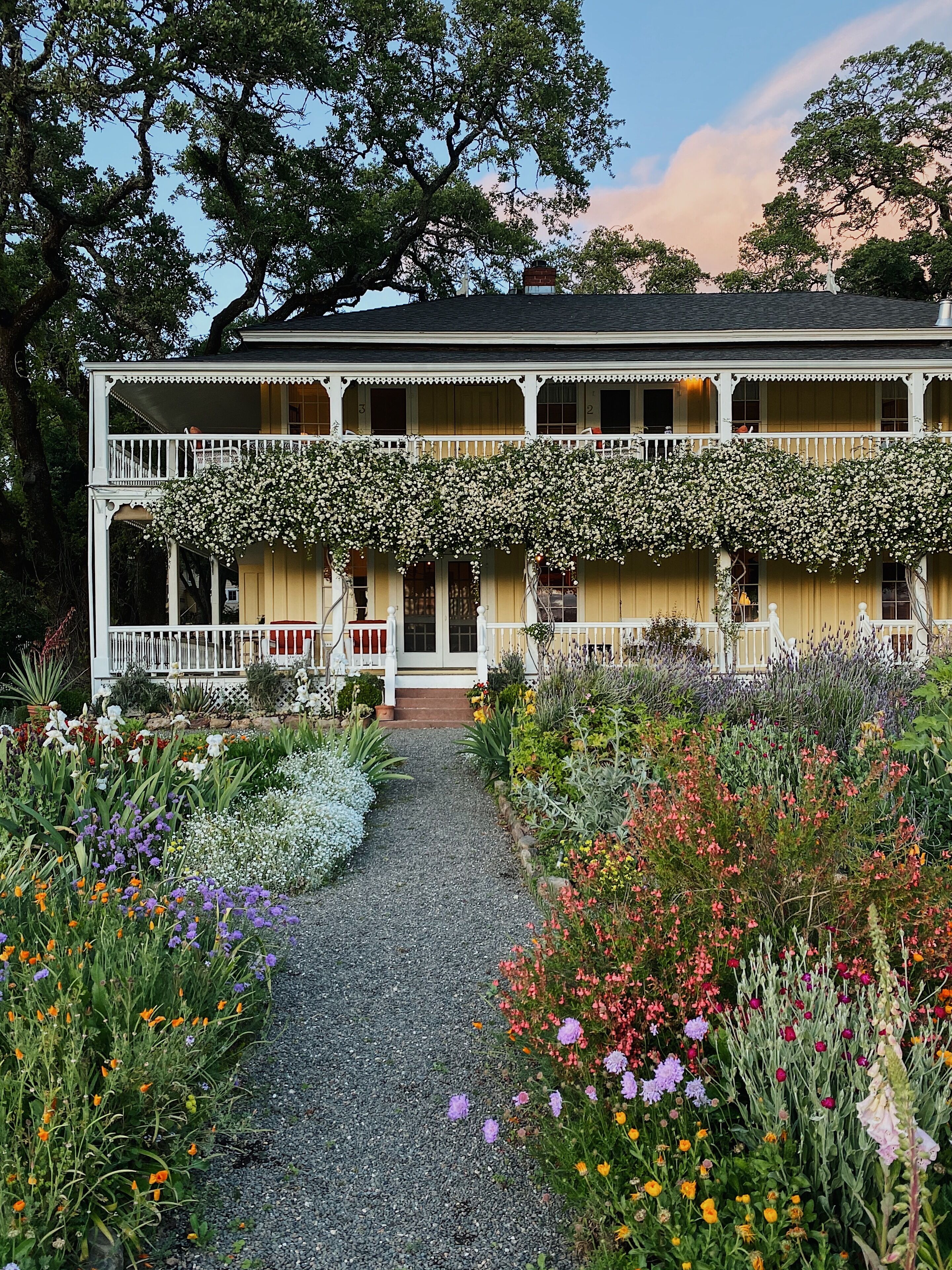 Uma casa de dois andares com varanda coberta e um jardim cheio de flores diversas.
