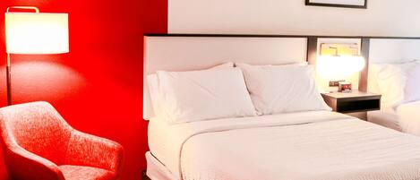 Quarto de hotel com uma cama, uma cadeira, uma mesa de cabeceira com abajur e um quadro na parede.