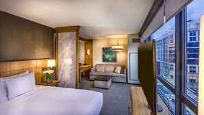 Quarto de hotel com uma cama grande, um sofá, uma escrivaninha e vista da cidade através de amplas janelas.