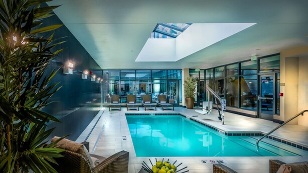 Uma área de piscina interior moderna com espreguiçadeiras, um painel de vidro e um claraboia.