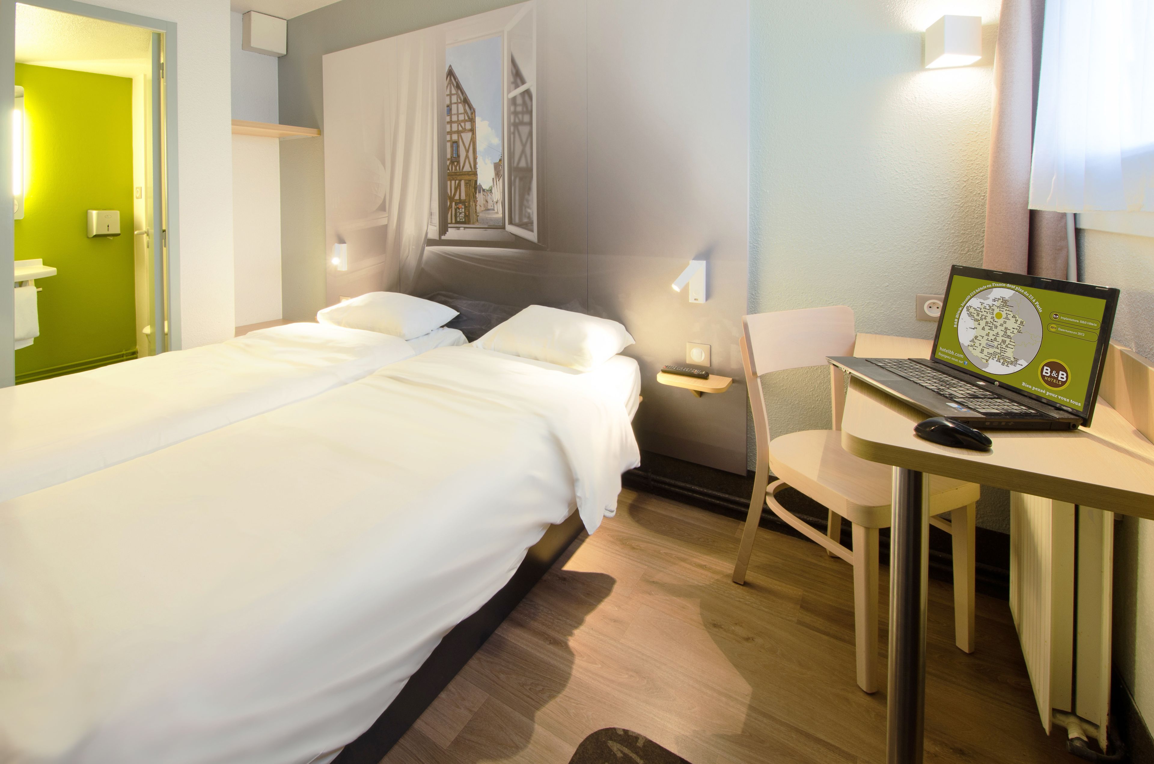 Quarto de hotel com cama, escrivaninha, um laptop e uma cadeira.