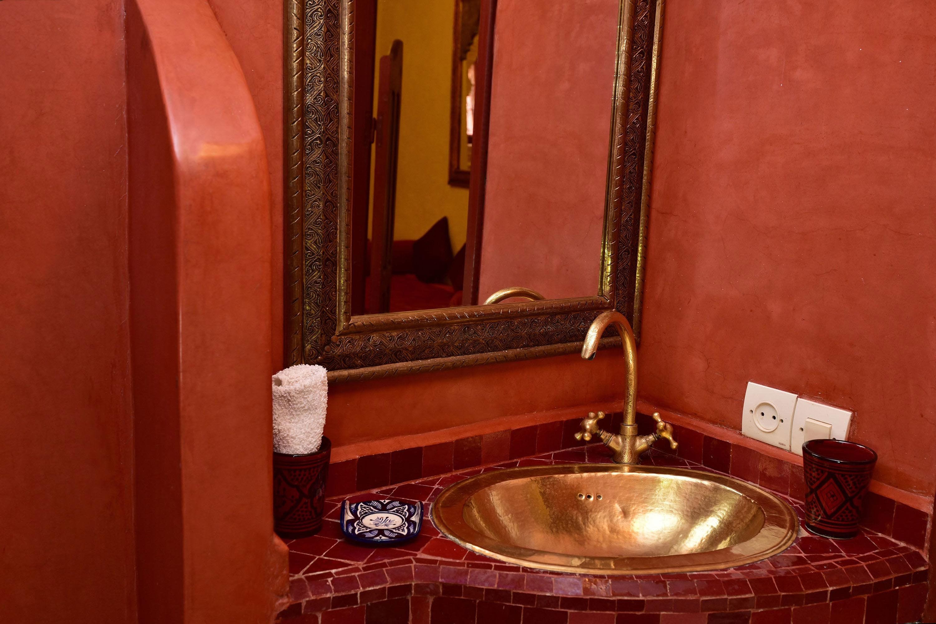 Casa de banho com lavatório dourado, espelho e parede revestida a azulejos vermelhos.