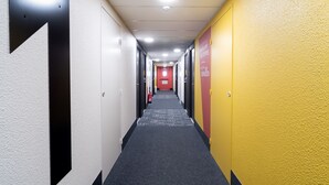 Um corredor com uma placa de seta preta à esquerda, uma porta vermelha no final e uma parede amarela à direita.