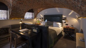 Quarto com teto de tijolos, cama com cabeceira, mesas de cabeceira com luminárias, escrivaninha com cadeira e janela com cortinas.