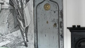 Um quarto com um penteado verde, uma parede branca com um mural em preto e branco, uma porta branca com fechadura de latão e uma lareira preta.