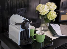 Uma cafeteira sobre um prato preto, com uma chávena verde e um vaso de flores brancas.