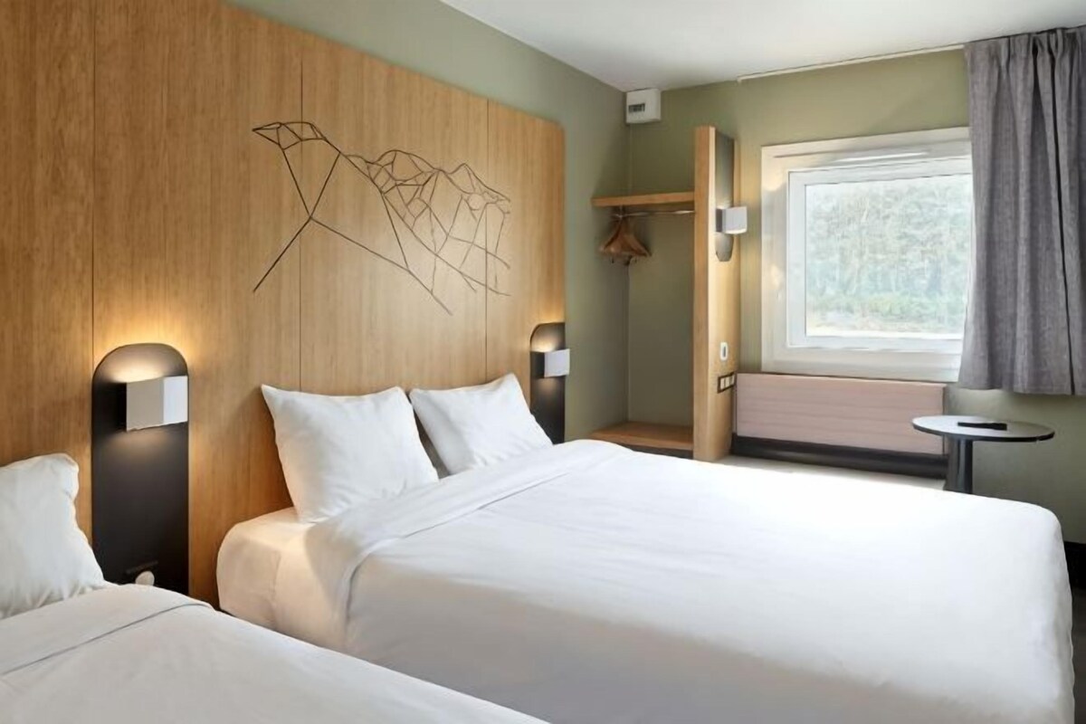 Quarto de hotel com duas camas, cabeceira de madeira com design geométrico, abajures, janela com cortinas e uma mesinha.