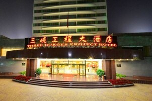 Entrada de hotel com placa em língua estrangeira, fachada bem iluminada e área ajardinada com degraus que levam à entrada.