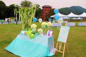 Montagem para evento ao ar livre com uma mesa coberta por um tecido azul claro, decorada com flores brancas e balões, e um suporte com uma lousa branca.