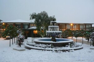 Área externa coberta de neve com uma fonte circular, um edifício iluminado e neve nas árvores.