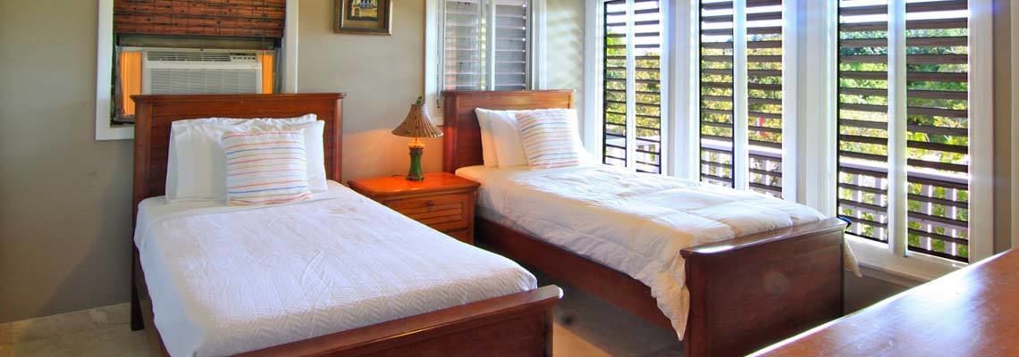 Quarto com duas camas de solteiro, mesa de cabeceira com abajur e janela com persianas.