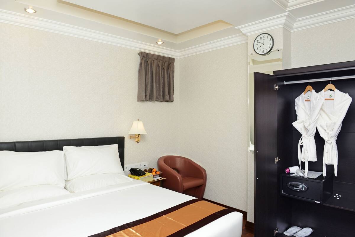 Quarto de hotel com uma cama, uma cadeira, um cabideiro e um relógio na parede.