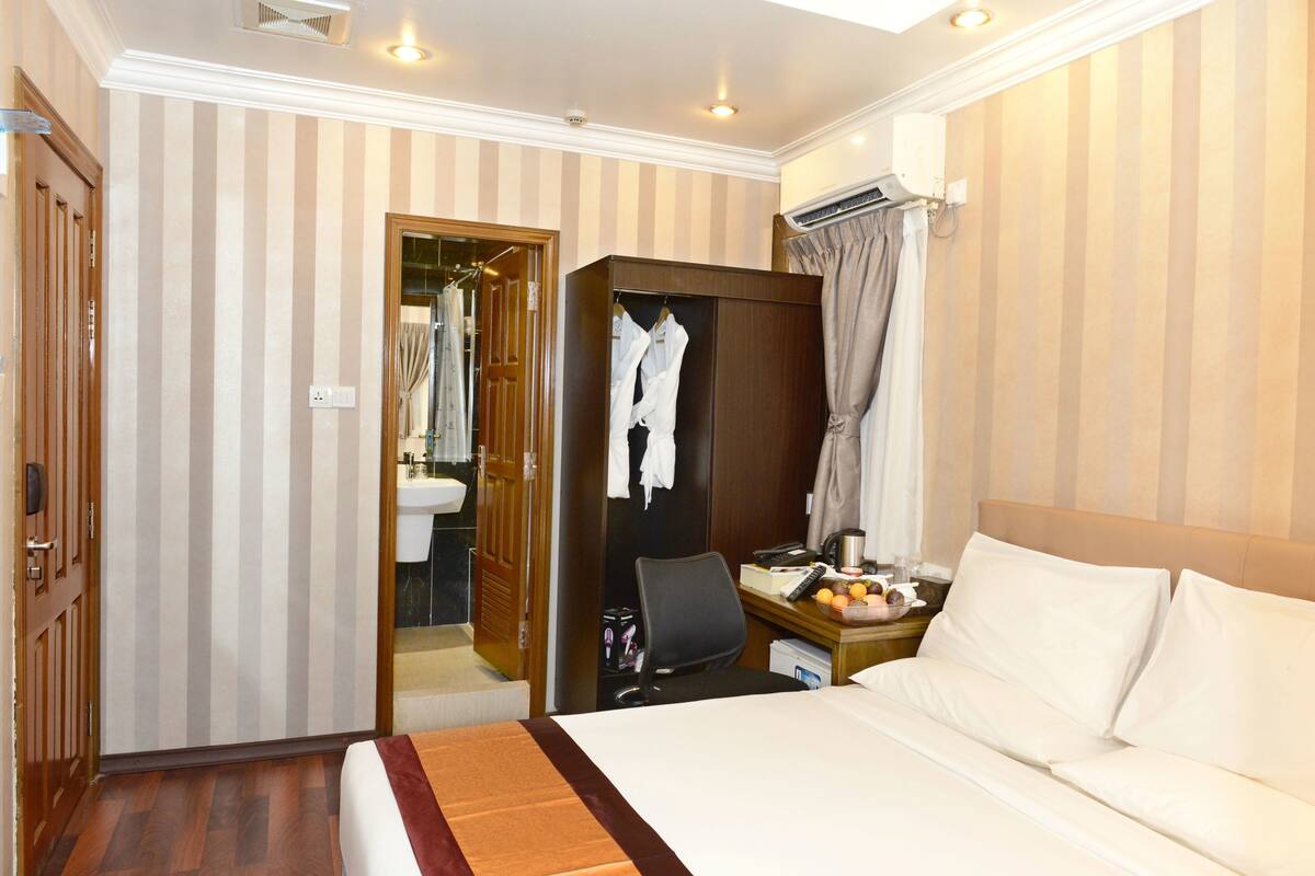 Quarto de hotel com cama, escrivaninha com cadeira, guarda-roupa e banheiro visível através de uma porta aberta.