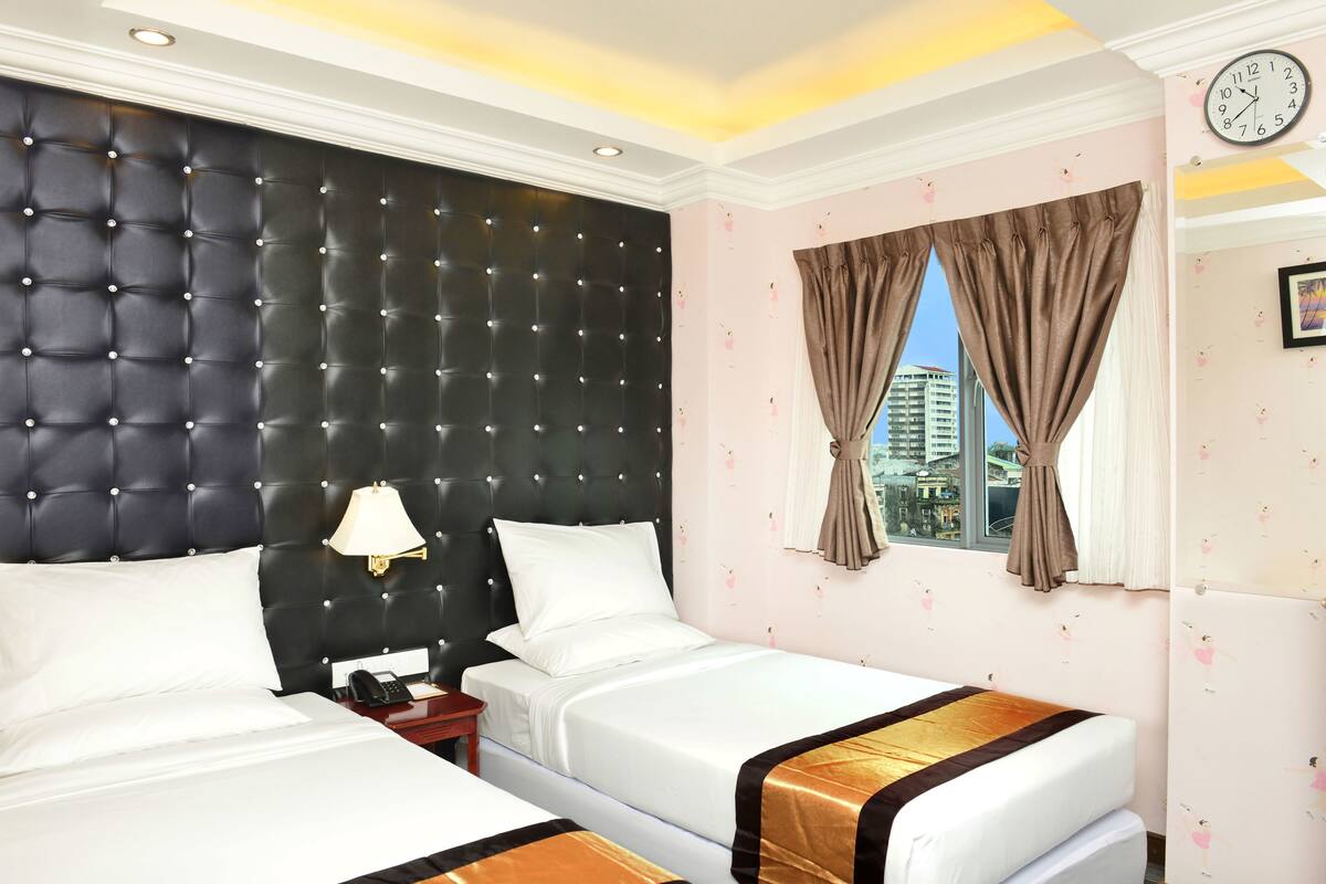 Quarto de hotel com uma cama grande, uma cama menor, uma mesa de cabeceira com luminária e uma janela com cortinas.