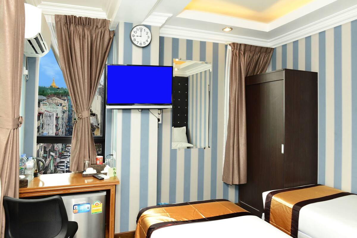 Quarto de hotel com duas camas, televisão, mesa com frigobar, relógio e vista para a paisagem urbana.