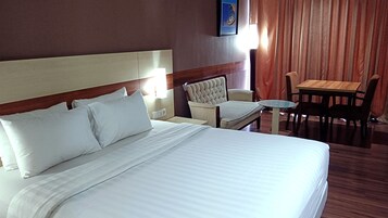 Quarto de hotel com uma cama grande, cabeceira de madeira, uma mesa redonda pequena, uma cadeira e uma luminária fixada na parede.