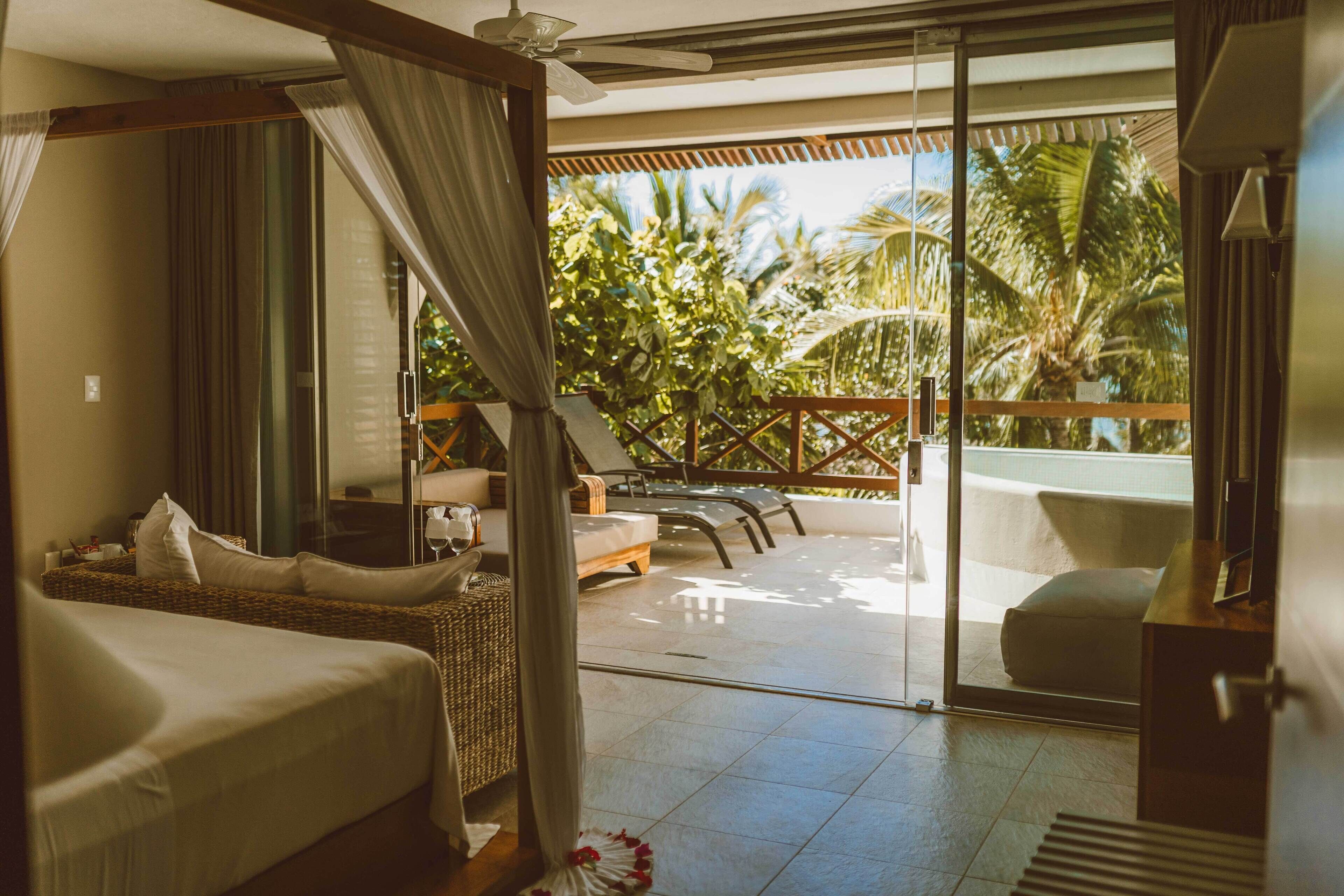 Quarto com cama, sofá, mesa de jantar e varanda com vista para palmeiras.