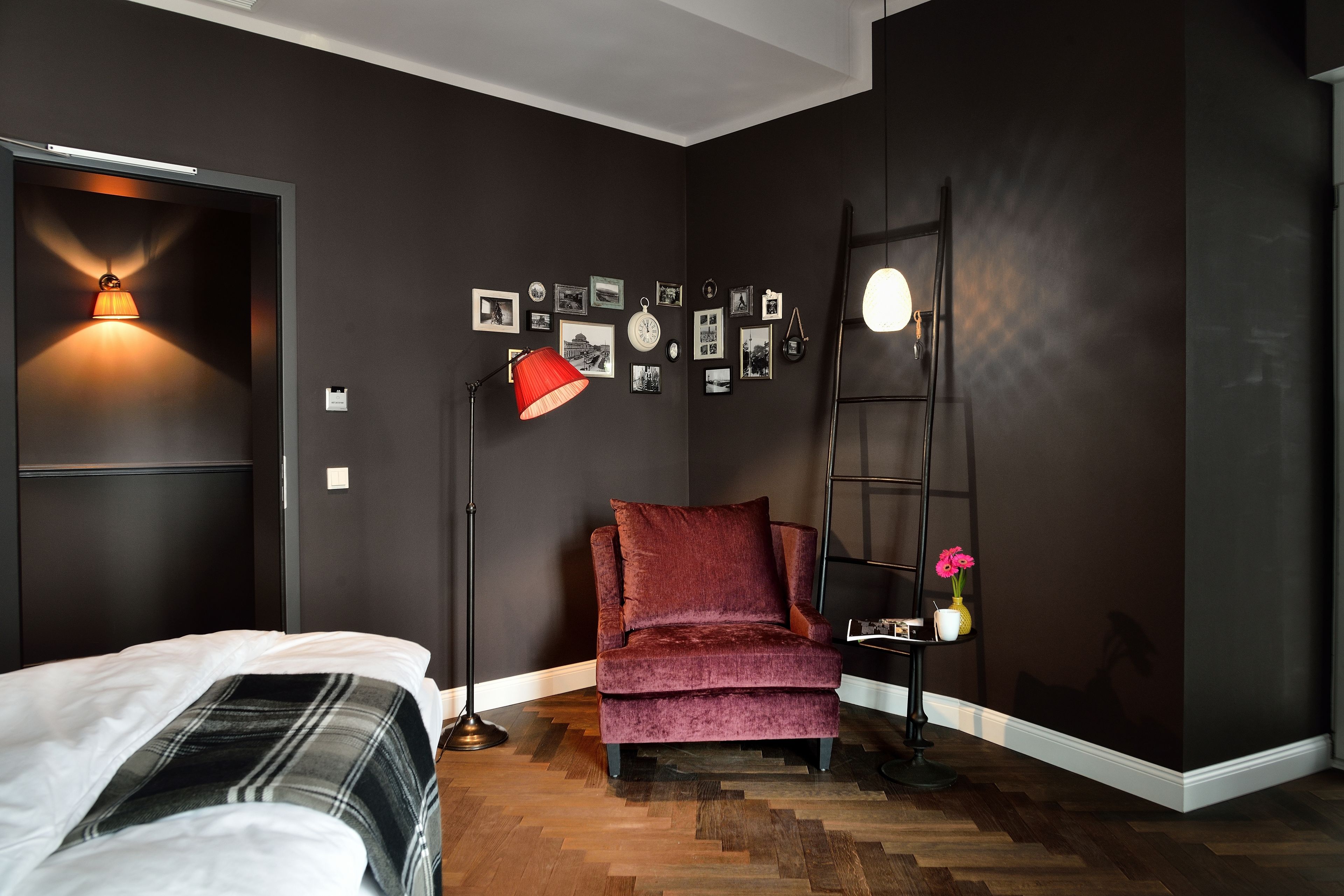 Quarto com decoração escura, uma cama, uma poltrona de veludo vermelho, um abajur e um painel de parede com molduras.