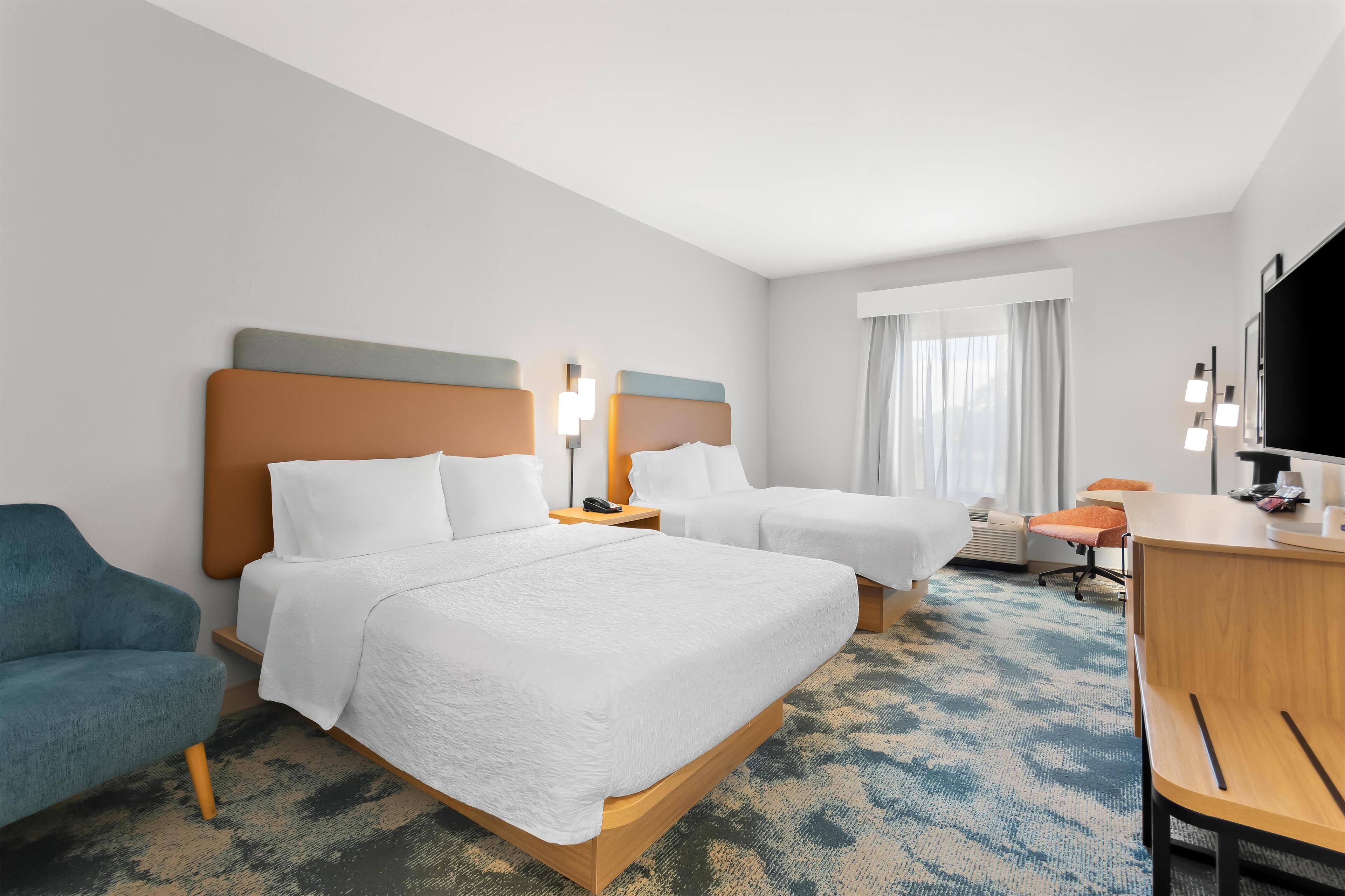 Quarto de hotel com duas camas, uma escrivaninha, uma cadeira e uma televisão.