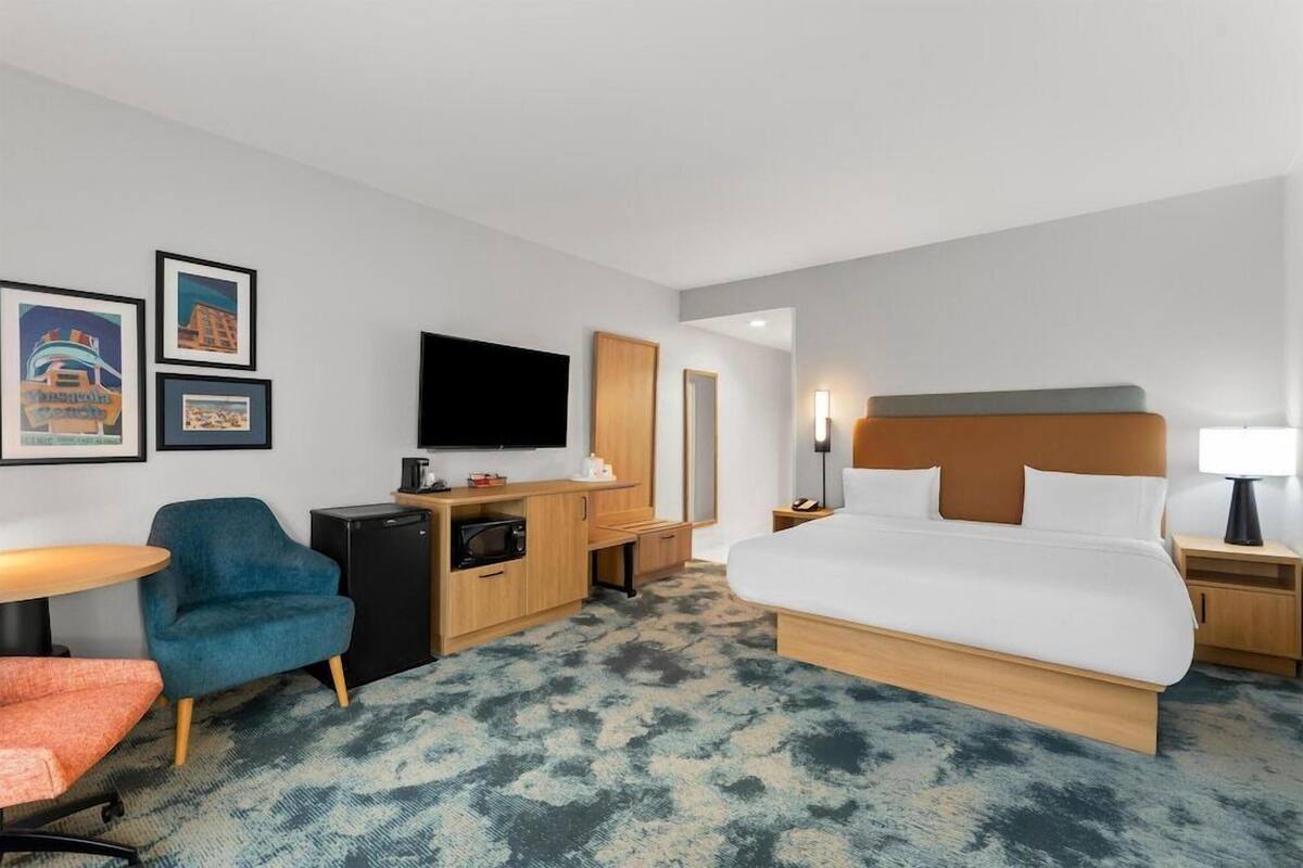 Quarto de hotel com cama, escrivaninha, cadeira, televisão e quadros na parede.