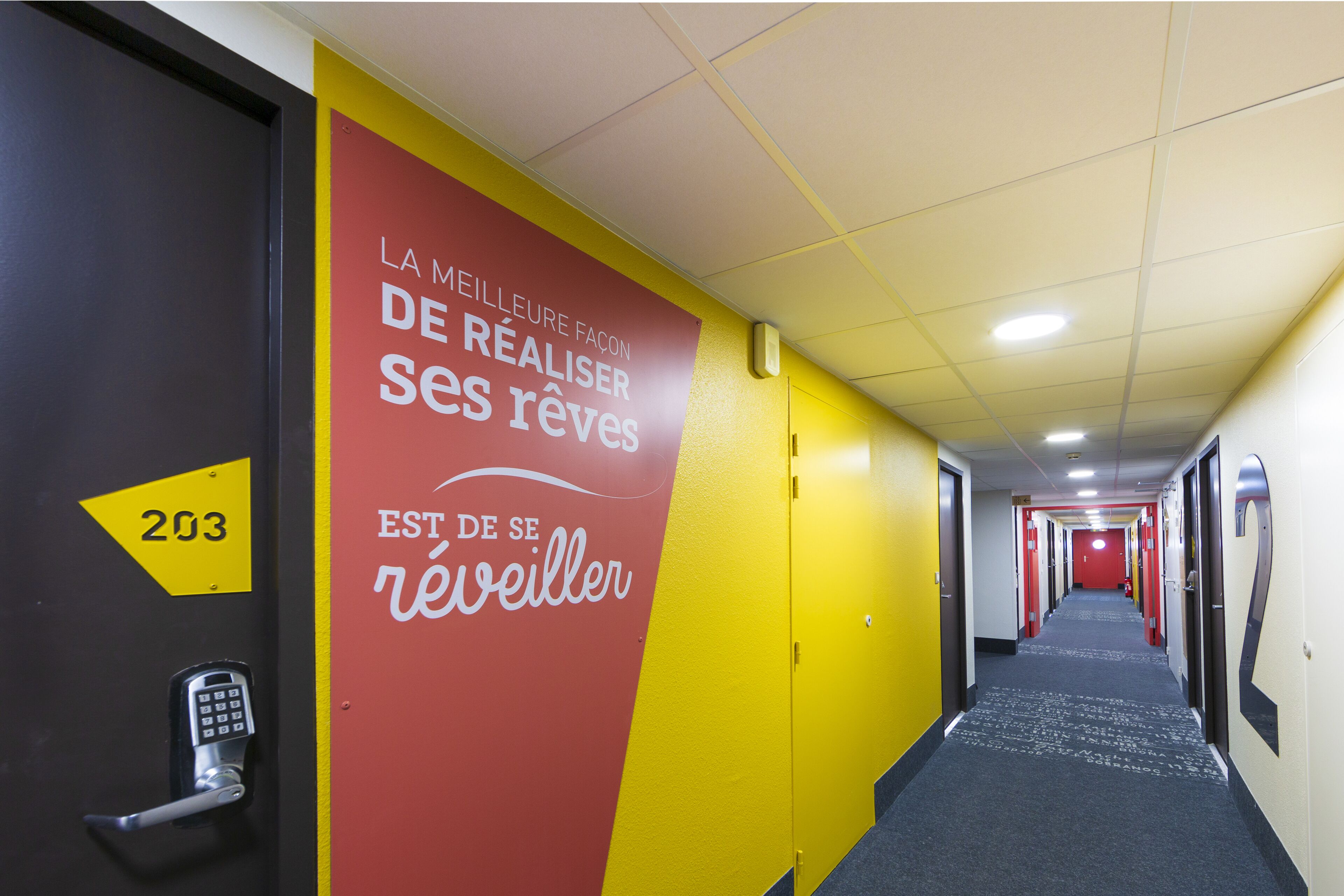 Corredor de hotel com parede em vermelho e amarelo, uma placa numérica e uma porta com fechadura eletrônica.