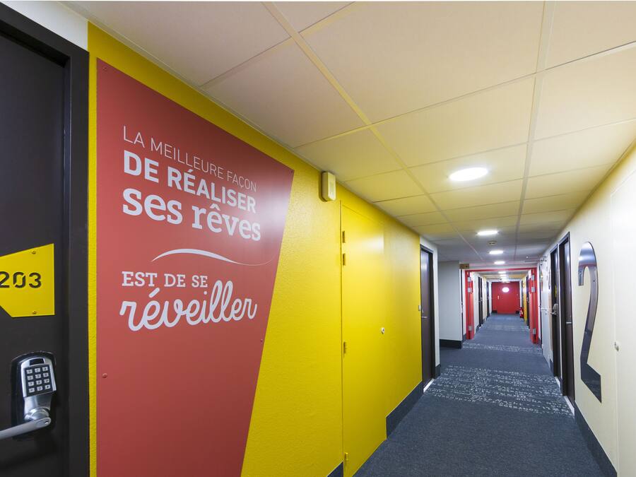 Corredor de hotel com parede em vermelho e amarelo, uma placa numérica e uma porta com fechadura eletrônica.