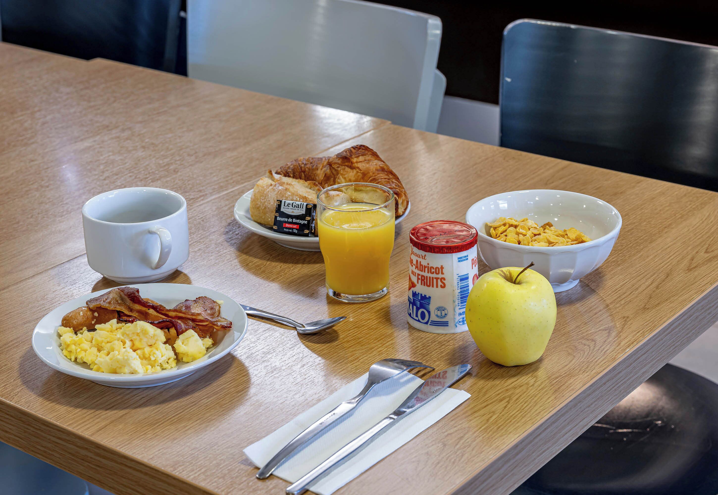 Uma mesa de café da manhã com pratos de ovos, bacon, torradas e um croissant. Há um copo de suco de laranja, uma tigela de cereal e uma maçã amarela.