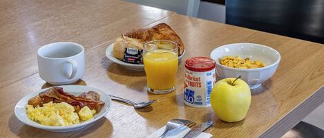 Uma mesa de café da manhã com pratos de ovos, bacon, torradas e um croissant. Há um copo de suco de laranja, uma tigela de cereal e uma maçã amarela.