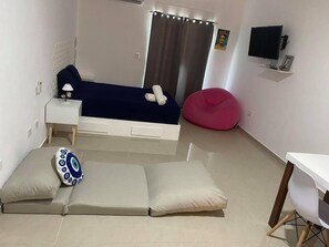Quarto pequeno e moderno com cama, mesa de cabeceira com luminária, poltrona de pelúcia rosa e televisão na parede.