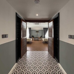 Corredor de hotel com piso estampado, área de estar com cadeiras e uma mesa com abajur.