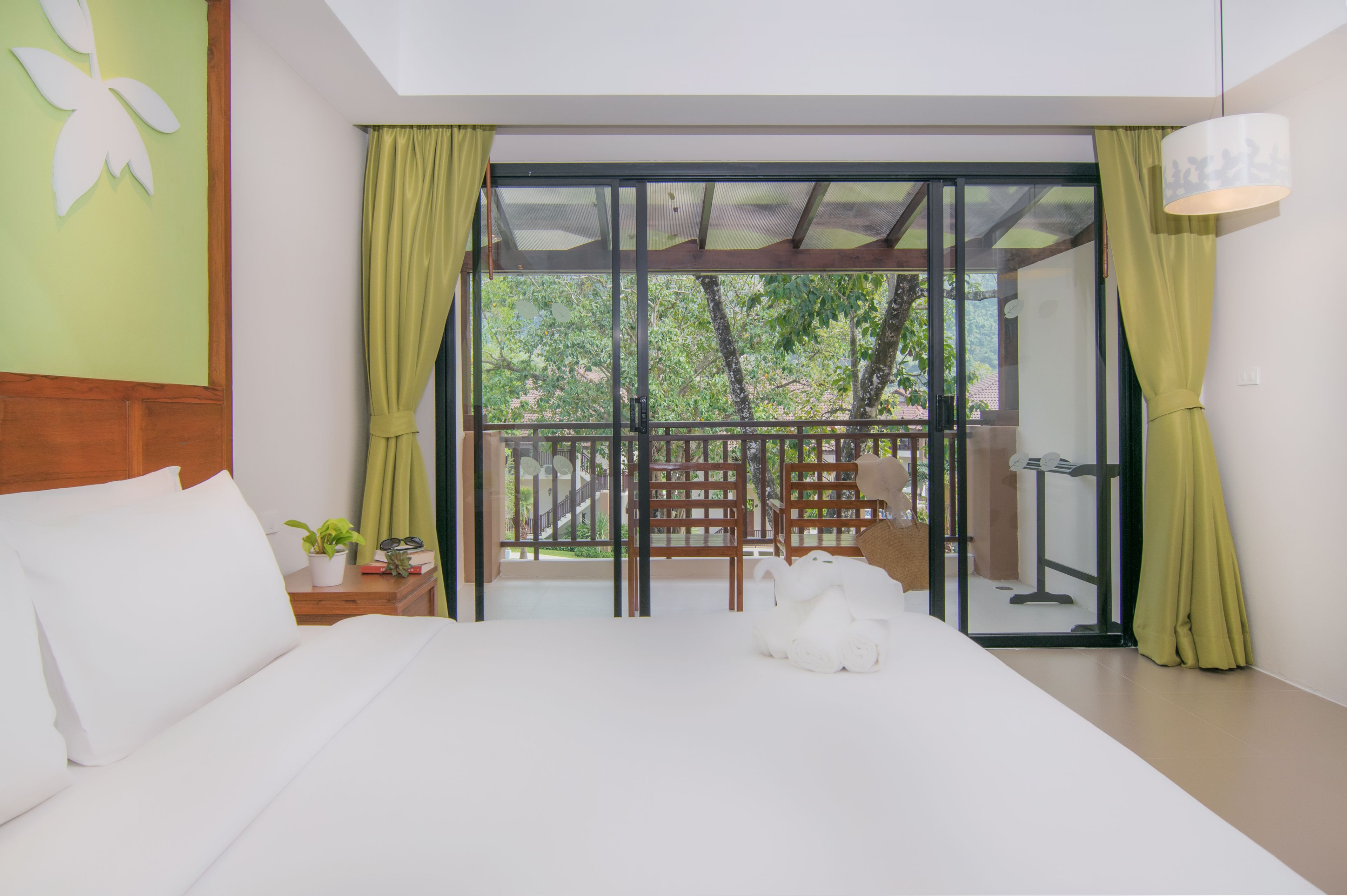 Quarto de hotel com uma cama grande, mesa de cabeceira com uma planta e vista para uma varanda com grades de madeira.