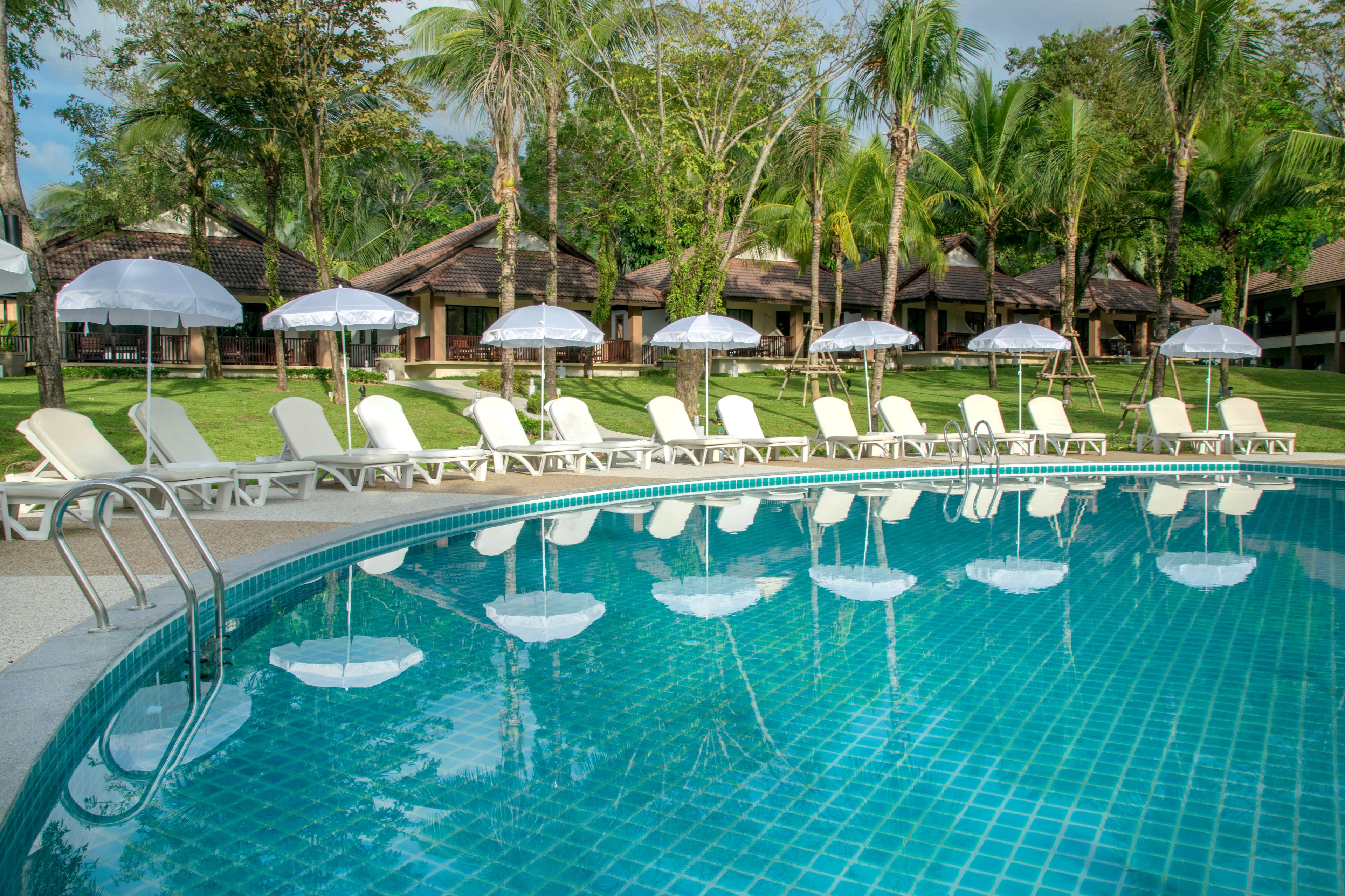 Piscina com cadeiras de sol brancas e guarda-sóis, cercada por palmeiras e edifícios de resort.