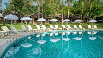 Piscina com cadeiras de sol brancas e guarda-sóis, cercada por palmeiras e edifícios de resort.