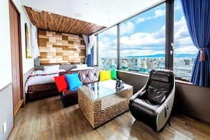 Quarto de hotel moderno com cama, mesa de centro de vidro, poltrona de massagem e uma grande janela com vista para a cidade.