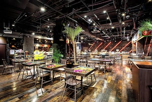 Um restaurante espaçoso com várias mesas e cadeiras, teto alto com canos aparentes e uma variedade de plantas.