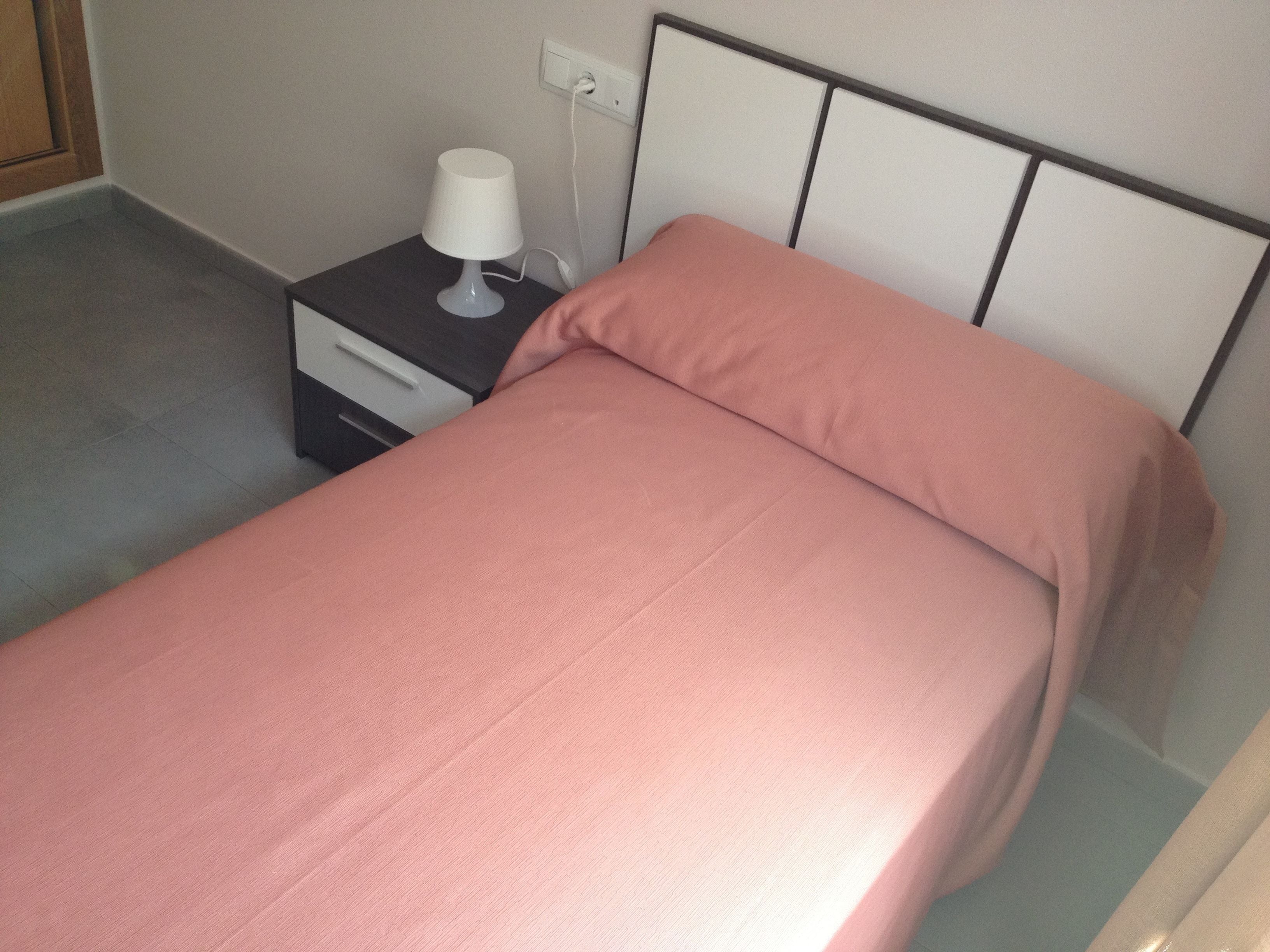 Uma cama pequena e bem arrumada, com colcha rosa e abajur branco sobre a mesa de cabeceira.