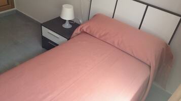 Uma cama pequena e bem arrumada, com colcha rosa e abajur branco sobre a mesa de cabeceira.