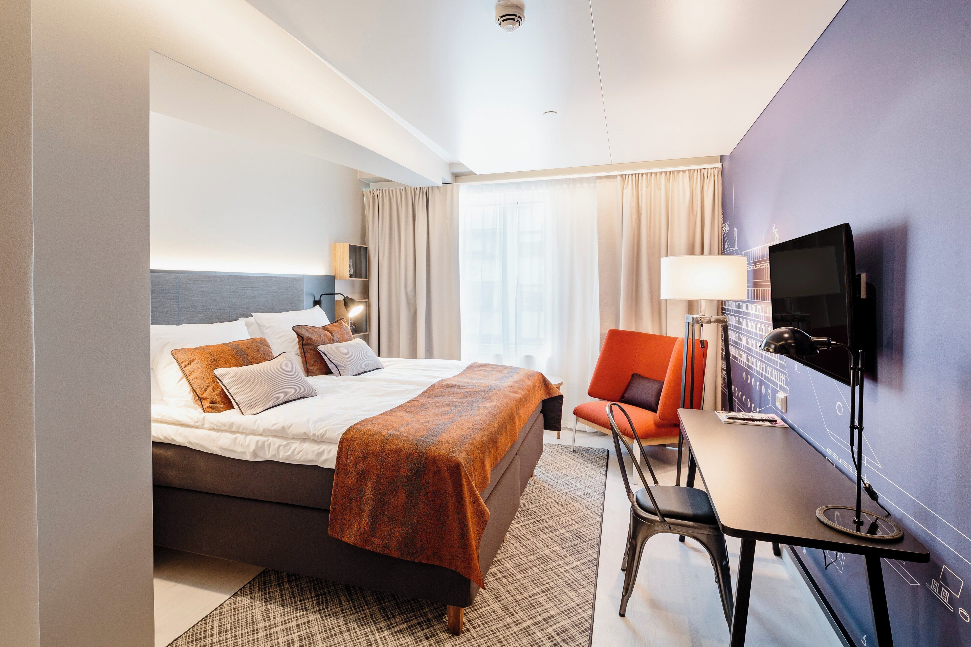Quarto de hotel com cama, escrivaninha, cadeira, televisão e janela com cortinas.